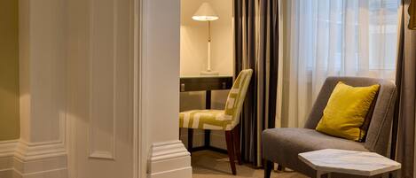 Quarto de hotel com escrivaninha, cadeira com almofada amarela, luminária e vista para o exterior através de cortinas translúcidas.