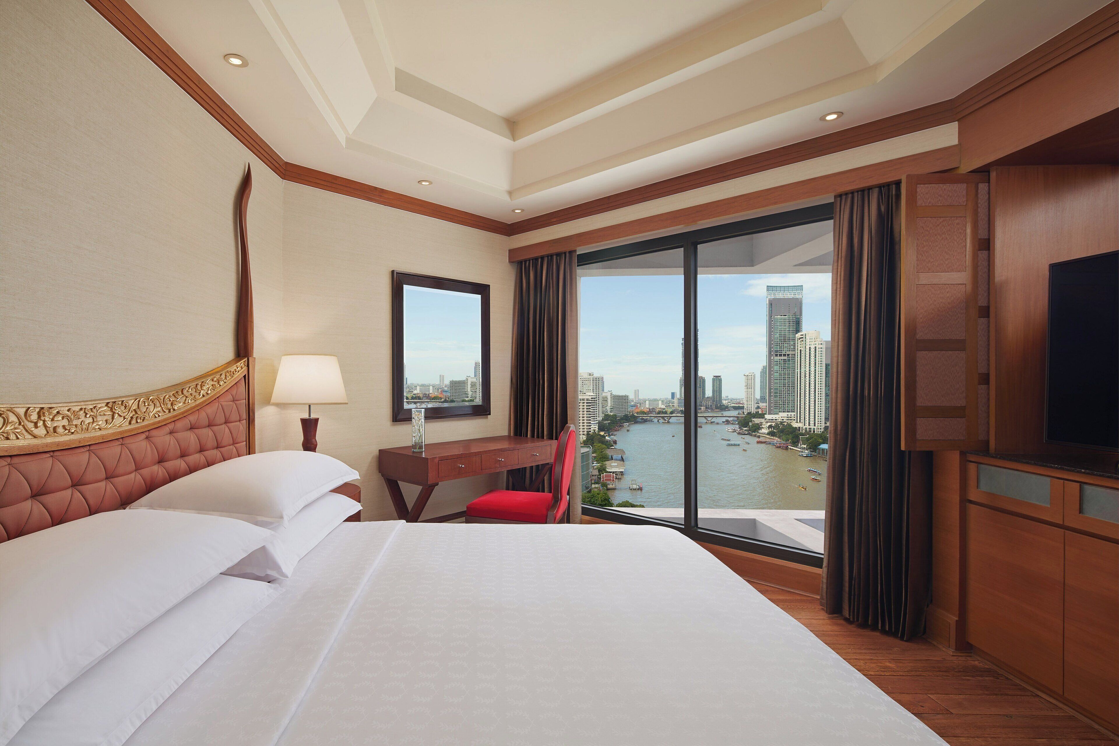 Quarto de hotel com uma cama grande, uma escrivaninha, uma cadeira, uma televisão e vista para a cidade e o rio.