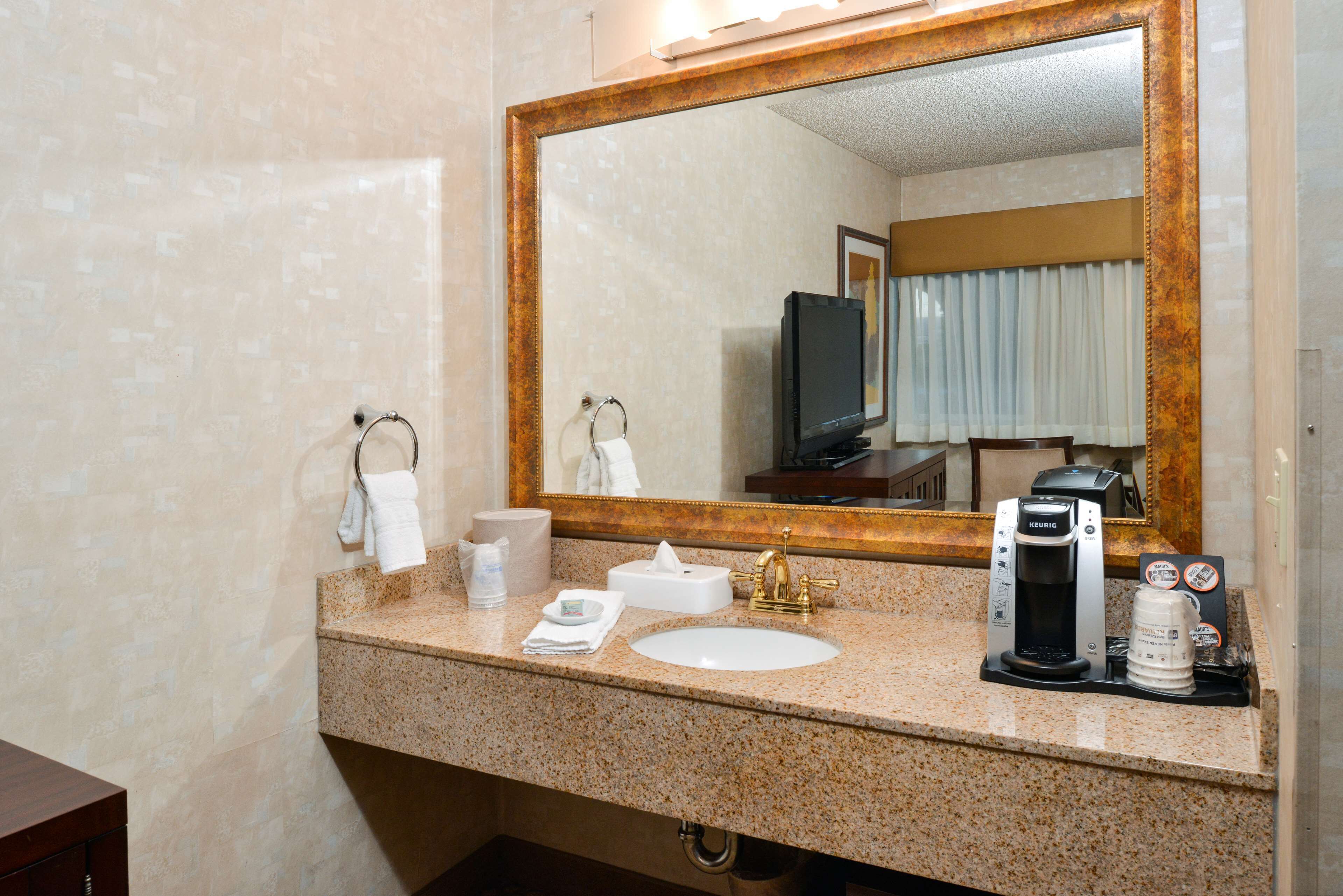Casa de banho de hotel com bancada de granito, lavatório, espelho, porta-toalhas, suporte de papel higiénico e máquina de café.
