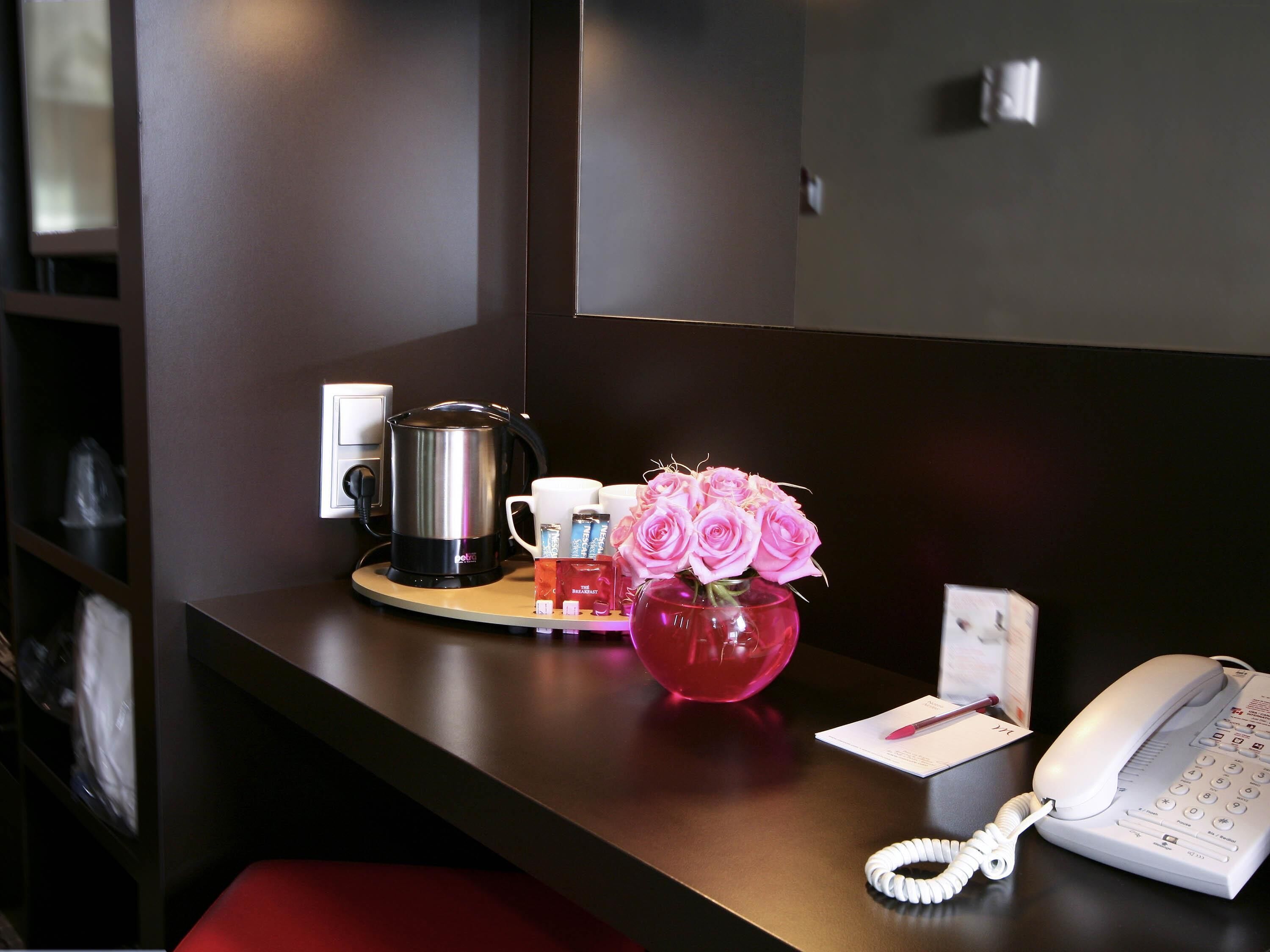 Balcão de recepção de hotel com uma cafeteira, um vaso de rosas cor-de-rosa, um telefone e um pequeno cartão no balcão.