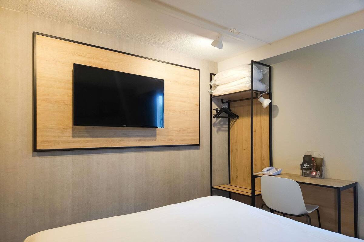 Quarto de hotel com cama, escrivaninha com cadeira, televisão de tela plana na parede, espelho e cabideiro.