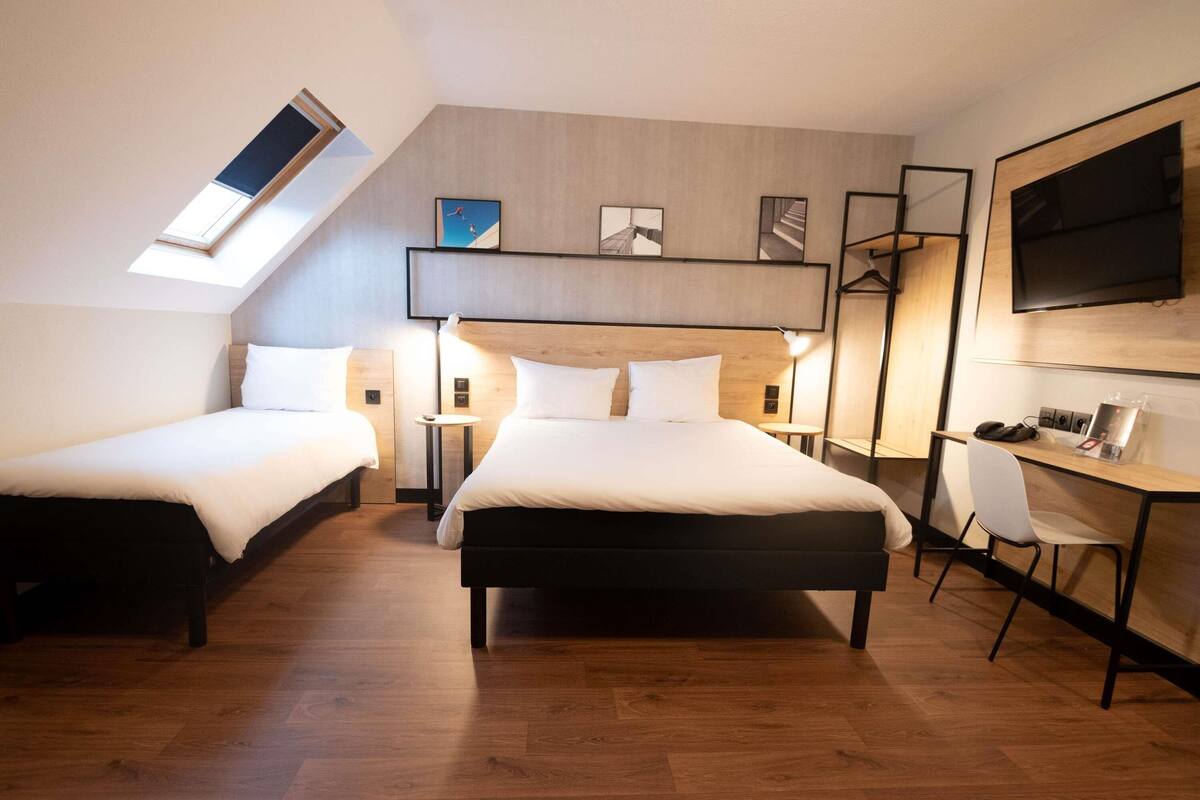 Quarto de hotel moderno com duas camas de solteiro, uma escrivaninha com cadeira, televisão na parede e um claraboia.
