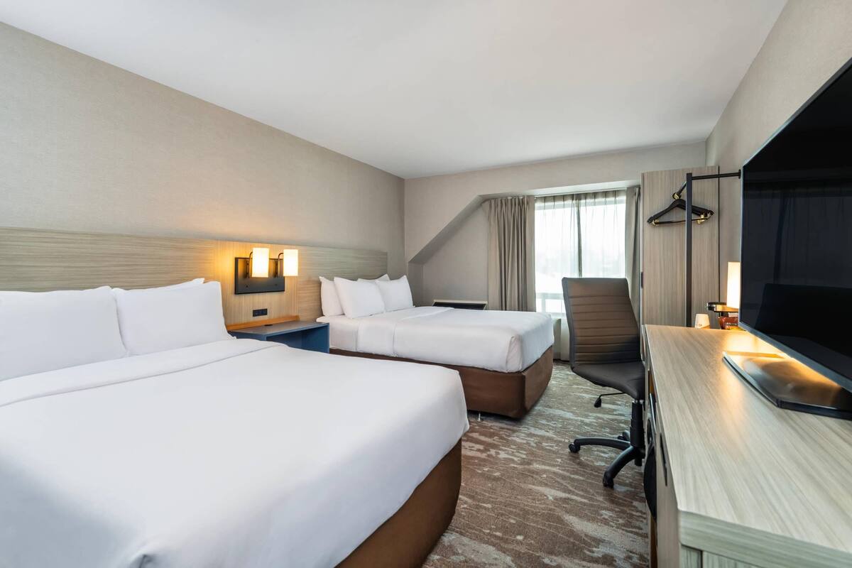 Quarto de hotel com duas camas, uma escrivaninha com televisão, uma cadeira e uma janela com cortinas.