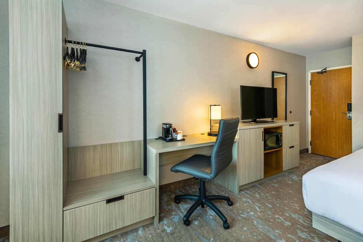 Quarto de hotel com cama, escrivaninha e cadeira, televisão, micro-ondas e guarda-roupa.