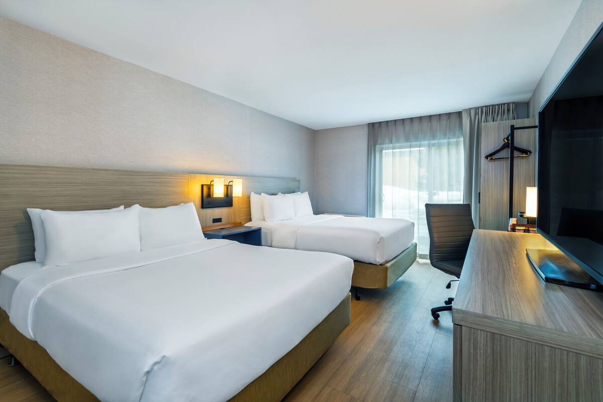 Quarto de hotel com duas camas, uma escrivaninha, televisão e janela com cortinas.