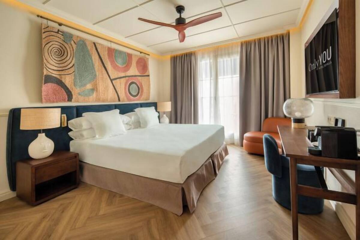 Quarto de hotel com cama, mesas de cabeceira, escrivaninha, cadeira, ventoinha no teto e uma grande pintura abstrata na parede.