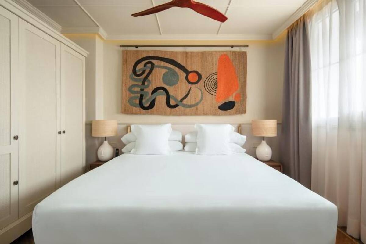Quarto de hotel com cama grande, duas luminárias laterais, cabeceira de madeira e uma obra de arte na parede.