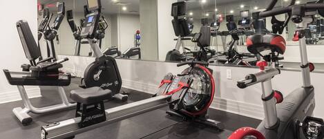 Academia com equipamentos de cardio, bancos de musculação e um espelho grande.