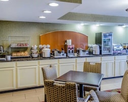Uma mesa de pequeno-almoço com máquina de café, torradeira e montra de comida. Cadeiras e uma mesa estão em primeiro plano.