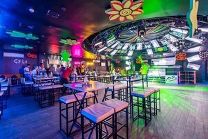 Um espaço de refeições interno vibrante, com várias mesas e cadeiras, um palco central com design circular e decorações coloridas no teto.