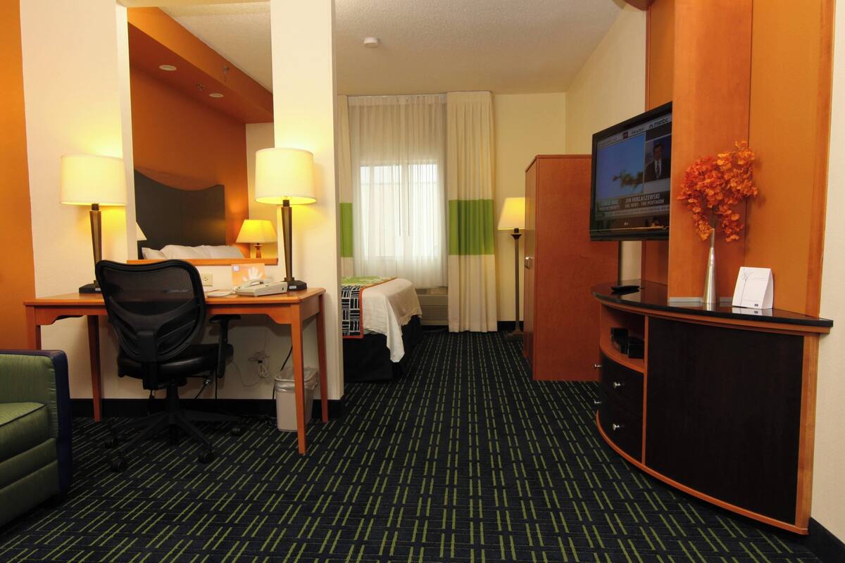 Quarto de hotel com escrivaninha, cadeira, cama, televisão e um pequeno armário.