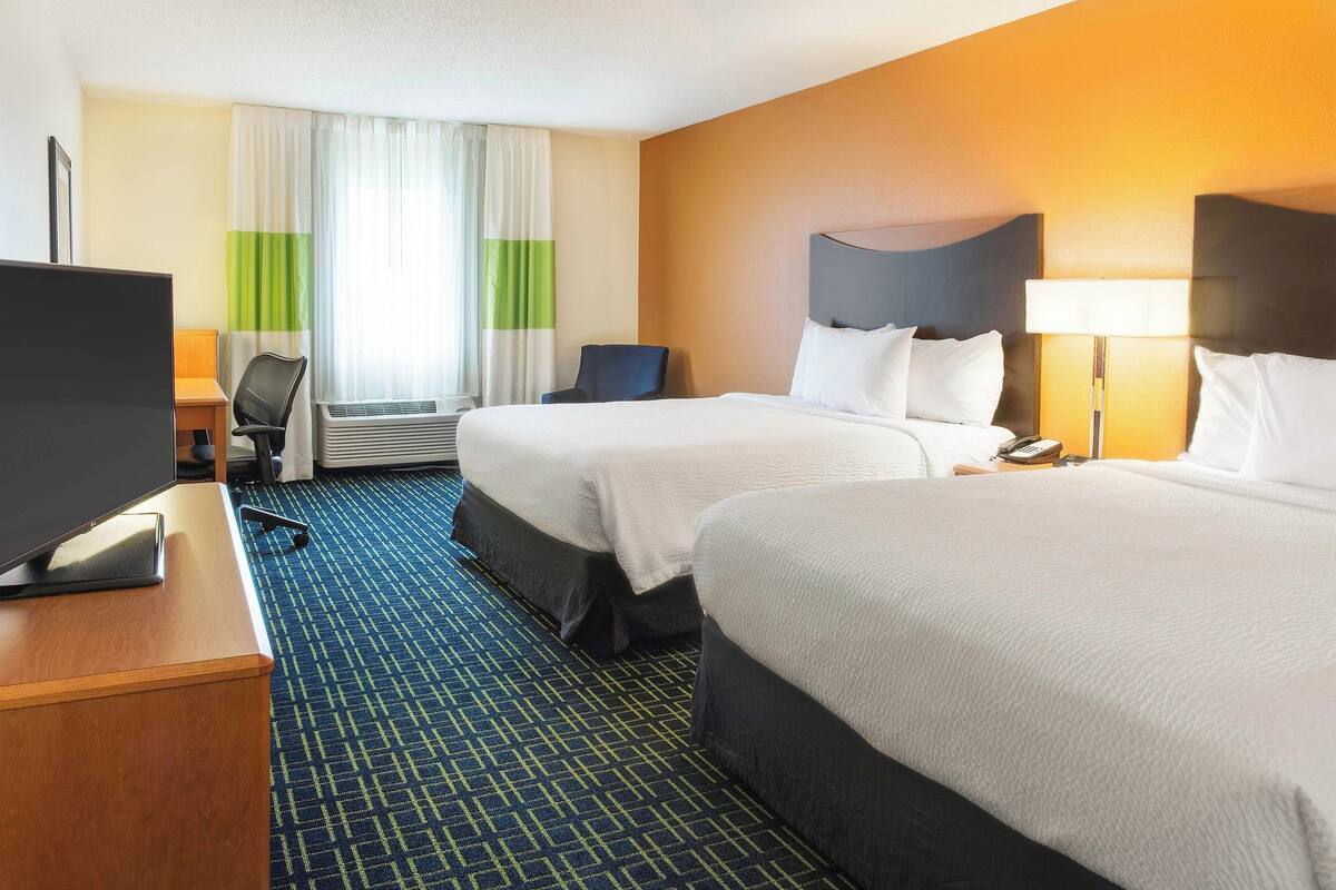 Quarto de hotel com duas camas, uma escrivaninha com computador, uma cadeira e uma televisão.
