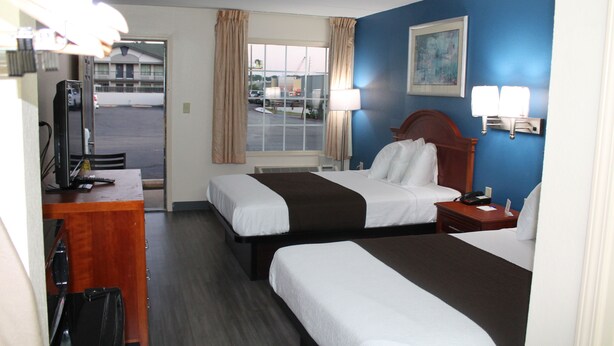 Quarto de hotel com duas camas, uma mesa com um computador, uma luminária e uma janela com cortinas.