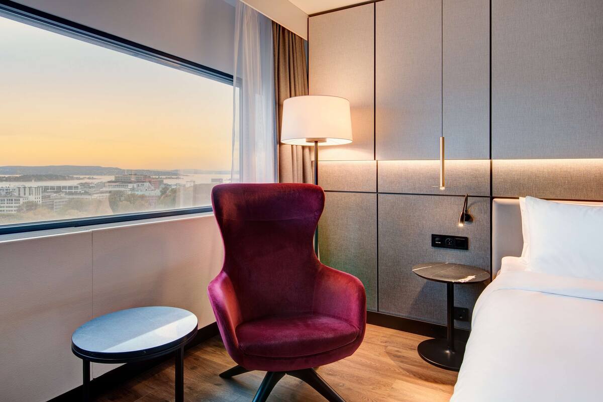 Quarto de hotel com uma janela grande, uma cadeira vermelha, uma mesa redonda pequena, um abajur e uma cama.