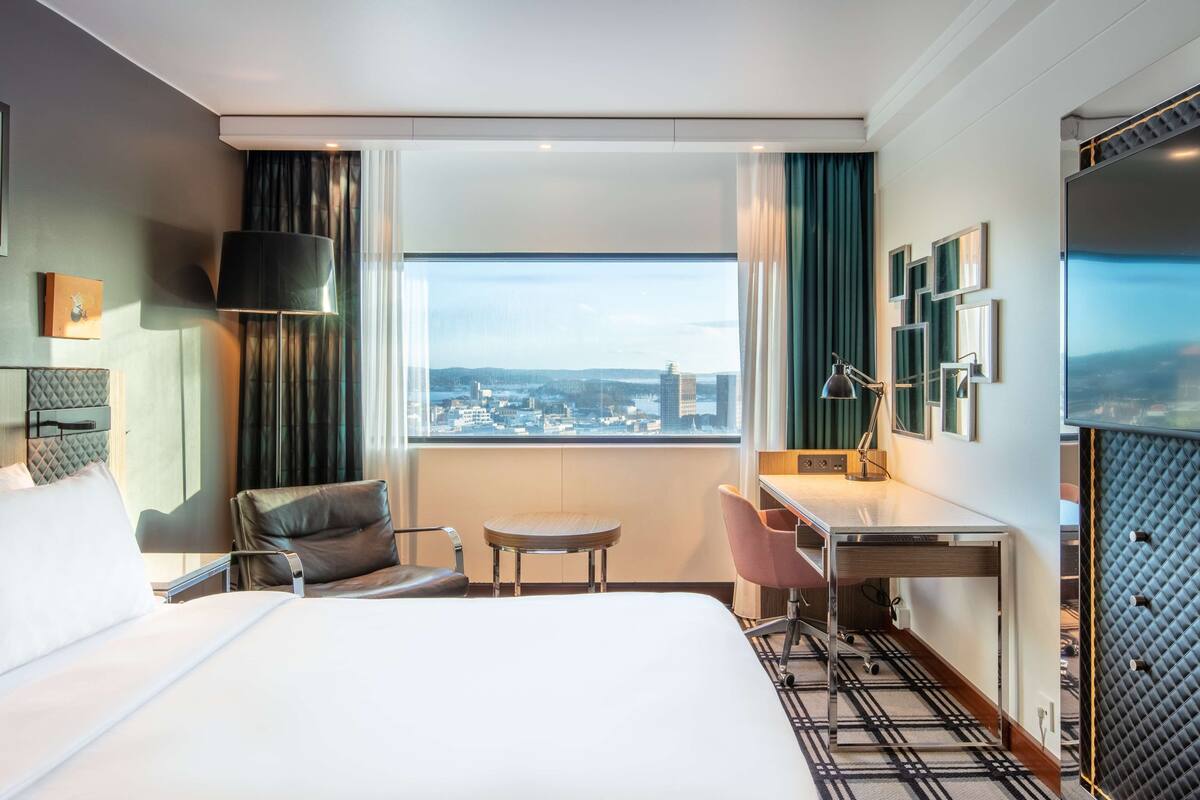 Quarto de hotel com uma cama grande, uma mesa, uma cadeira e vista para a cidade.