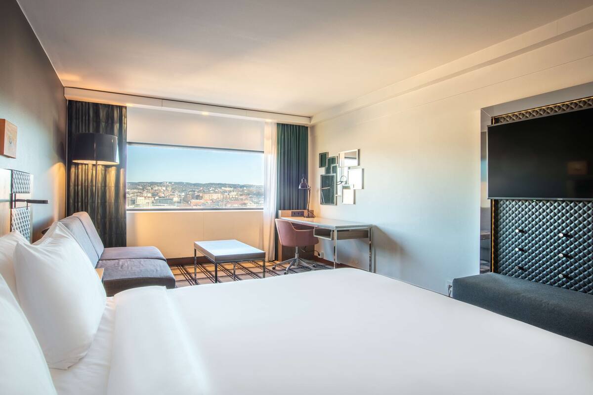 Quarto de hotel com uma cama grande, uma escrivaninha, uma cadeira, uma mesinha, um sofá, uma janela com vista para a cidade e uma televisão.
