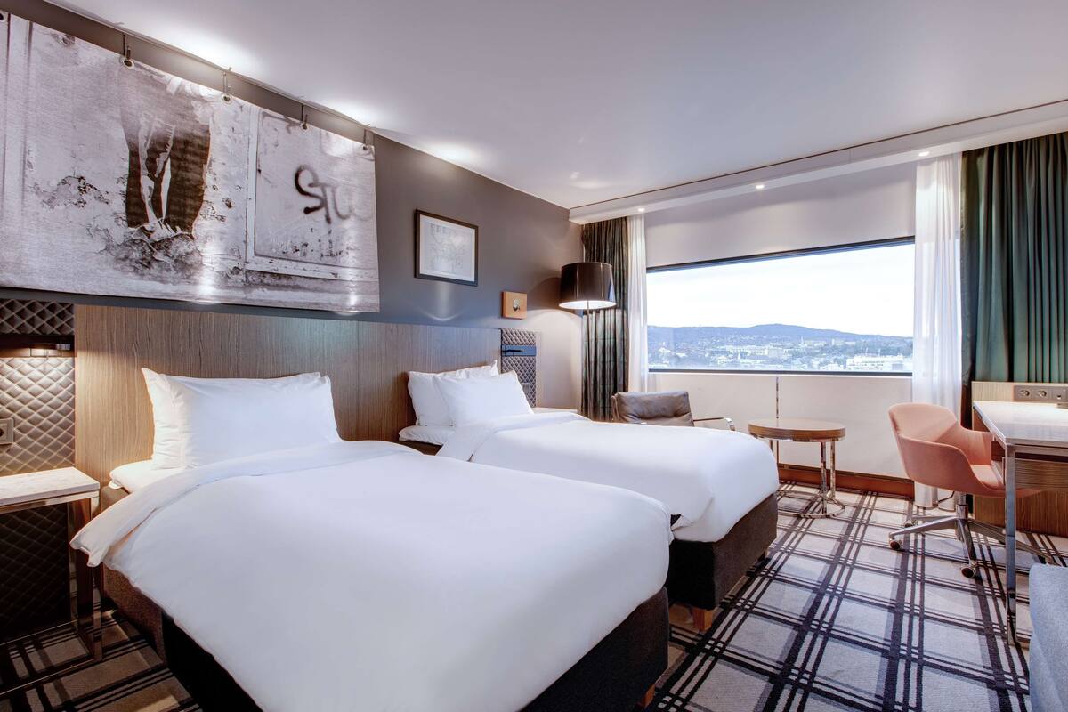 Quarto de hotel com duas camas, uma escrivaninha, uma cadeira, vista para a cidade e uma grande obra de arte na parede.