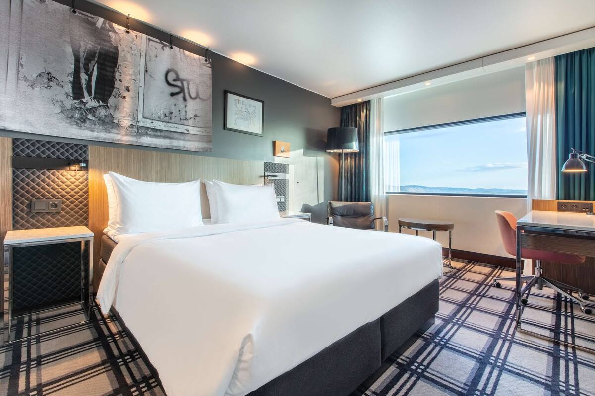 Quarto de hotel com uma cama grande, uma escrivaninha, uma cadeira, um abajur e vista para a cidade.