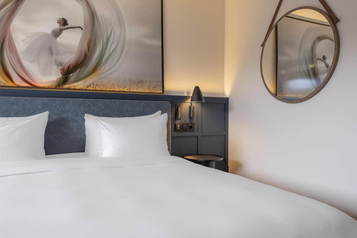 Quarto de hotel com uma cama grande, cabeceira, mesa de cabeceira com abajur e espelho fixado na parede.