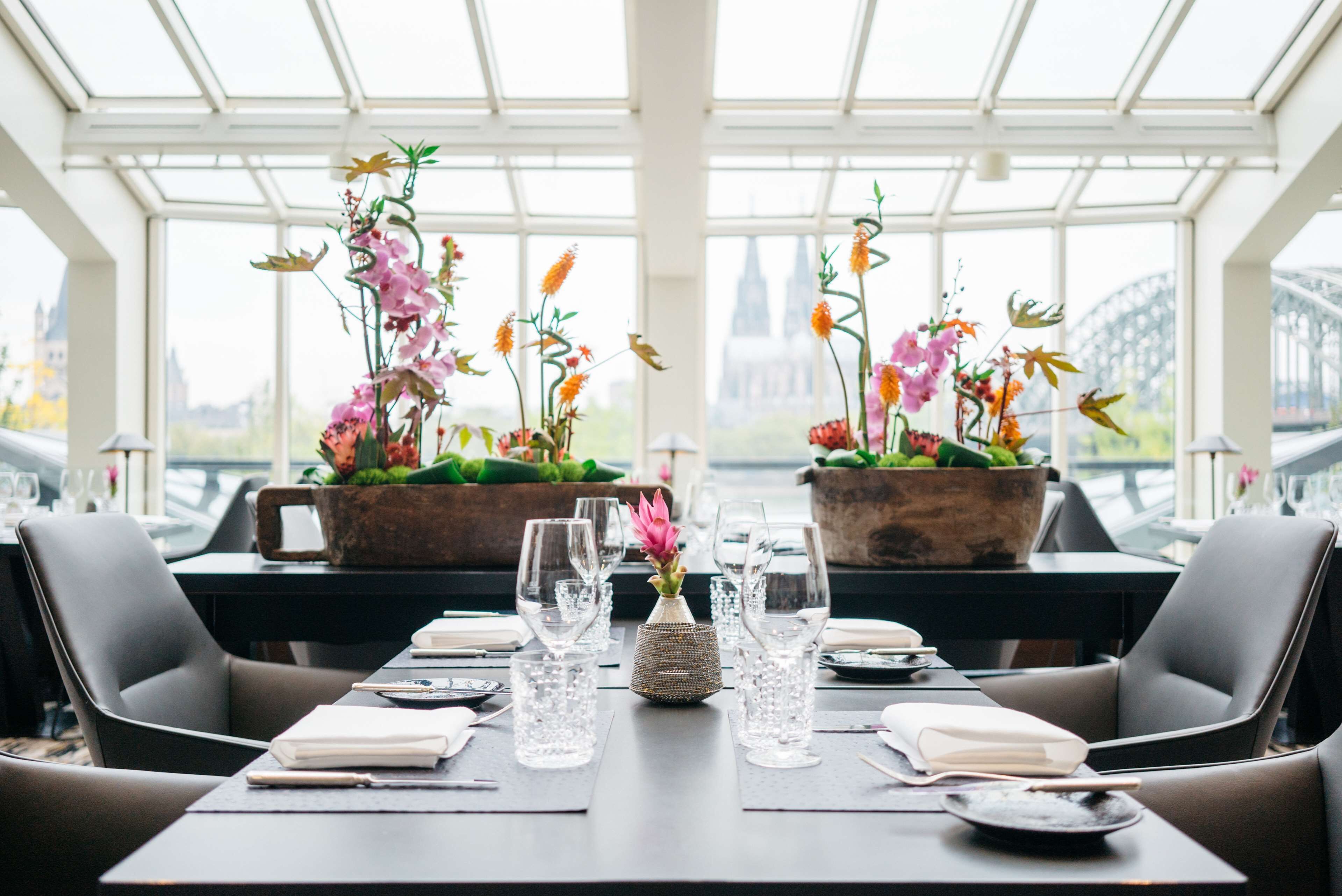 Mesa de restaurante posta com copos de vidro transparente, um arranjo de centro de mesa tecido e vista para um horizonte urbano através de grandes janelas.