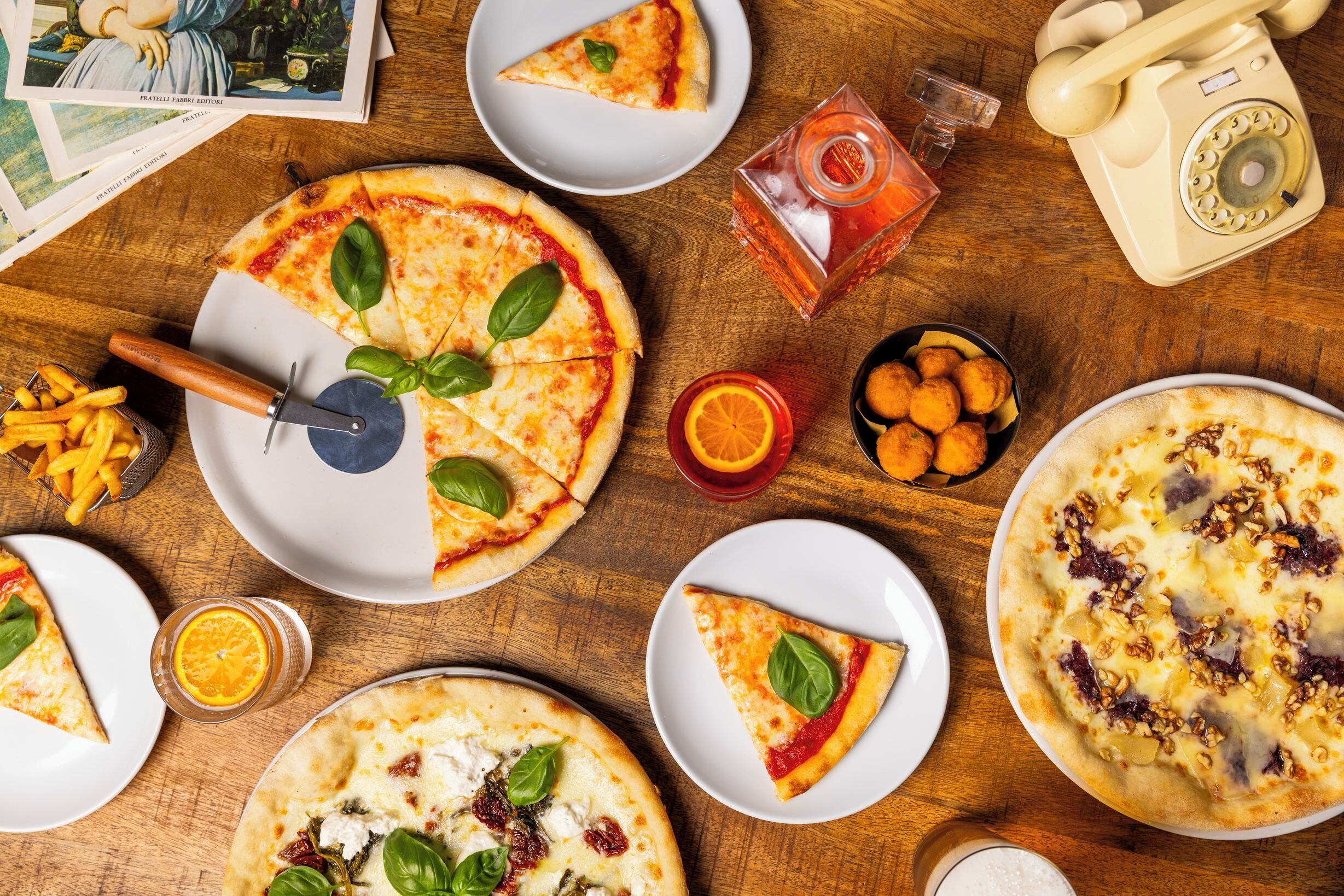 Uma mesa de madeira com várias pizzas, um cortador de pizza, um copo de suco de laranja, um copo de bebida vermelha, um recipiente com tater tots e um telefone antigo.