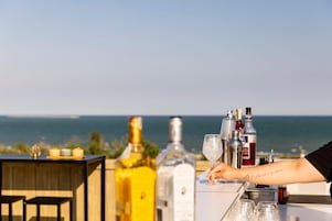 Uma área de bar com garrafas e copos, uma mesa de madeira e vista para o mar.