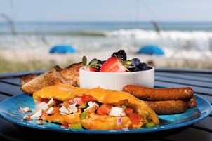 Prato com alimentos para o café da manhã, incluindo ovos, salsicha e torradas, com vista para a praia ao fundo.