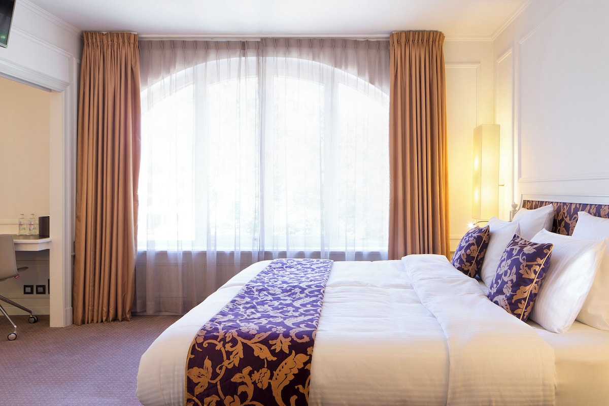 Quarto de hotel com uma cama grande, uma mesinha de cabeceira, um abajur e vista para o exterior através de cortinas finas.