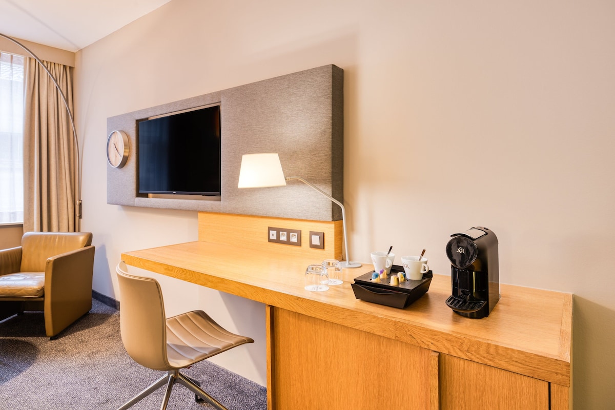 Quarto de hotel com escrivaninha de madeira, televisão na parede, luminária, cafeteira e uma cadeira.