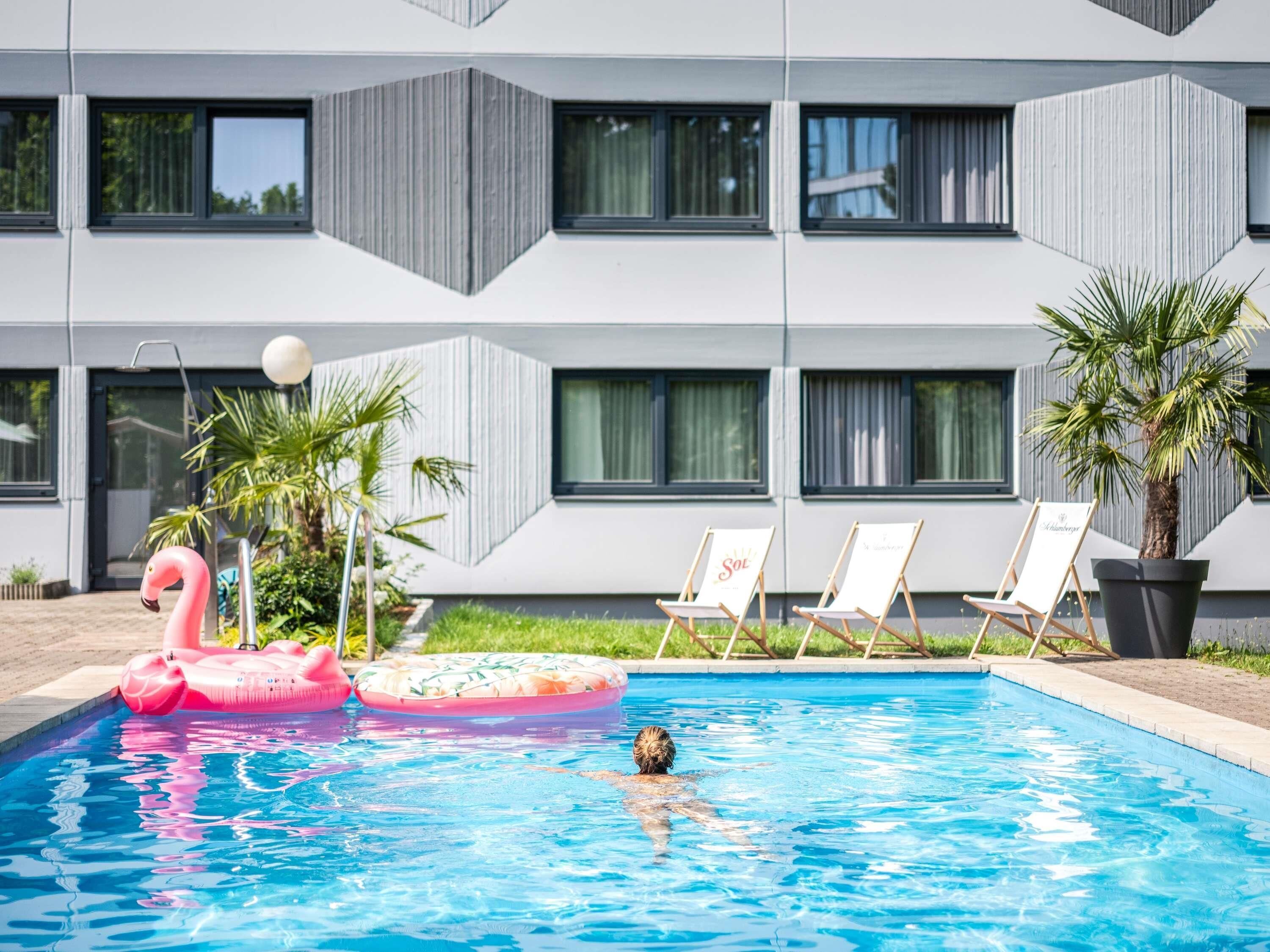 Piscina com um flutuador em formato de flamingo rosa e cadeiras de descanso em frente a um edifício moderno com grandes janelas.
