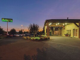 Entrada de hotel com placa da La Quinta Inn, estacionamento e um edifício com fachada de pedra.