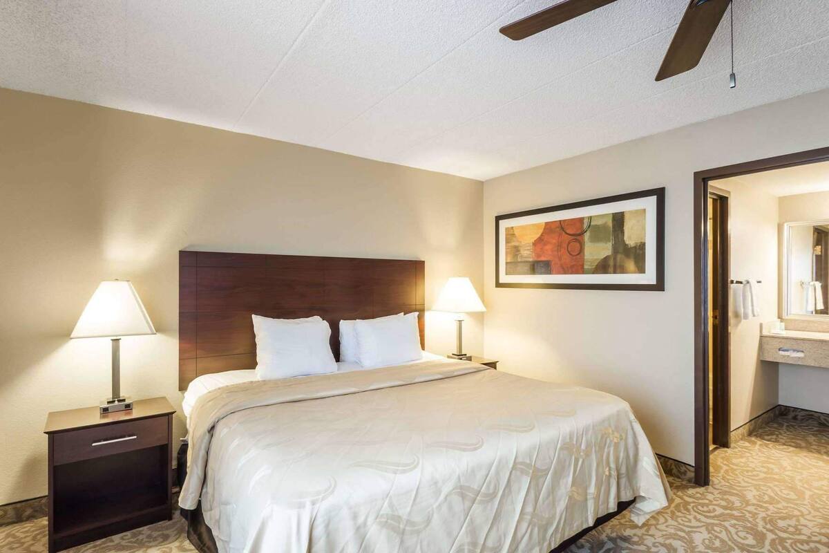 Quarto de hotel com cama, duas mesas de cabeceira com luminárias, um ventilador de teto, um quadro na parede e um banheiro visível por uma porta aberta.