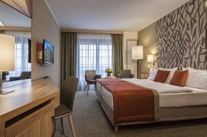Quarto de hotel com cama, secretária, cadeira e uma grande janela com cortinas.