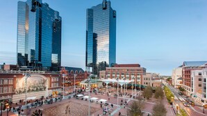 Uma praça com edifícios modernos, uma fonte e um monumento histórico.