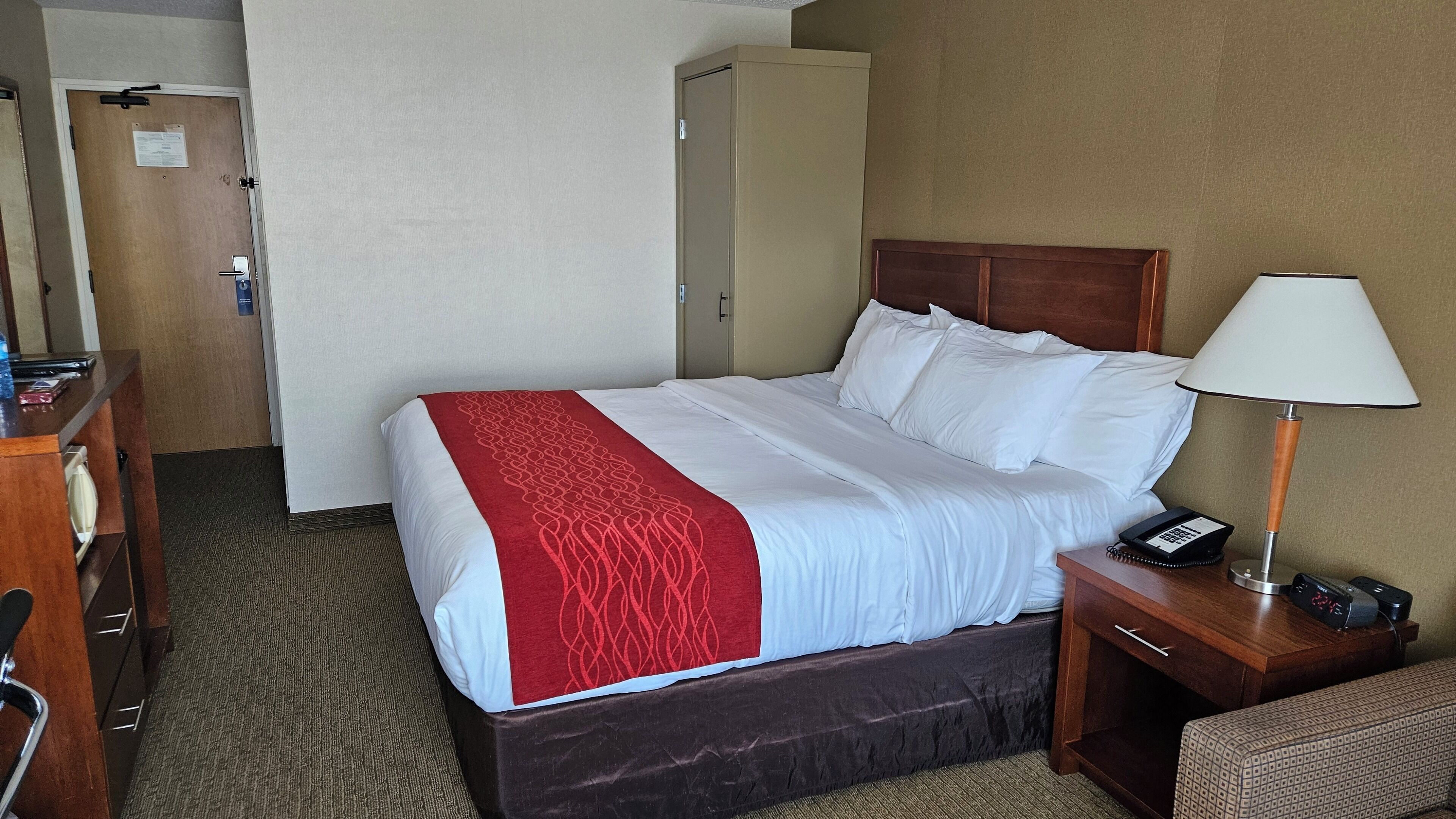 Quarto de hotel com cama, escrivaninha, cadeira, abajur e telefone.