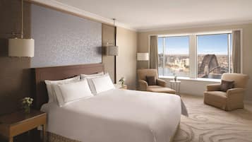 Quarto de hotel com uma cama grande, duas poltronas, uma mesinha e vista para a cidade através de amplas janelas.
