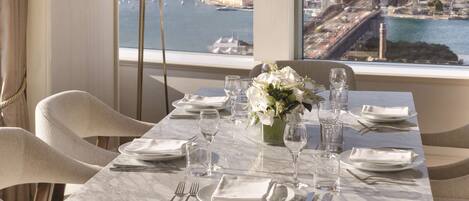 Uma área de jantar com uma mesa posta para quatro pessoas, um vaso de flores e vista para um horizonte urbano com uma ponte.