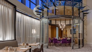 Um restaurante moderno com teto de vidro, área de refeições central com cadeiras roxas e piso com padrão de pedra.