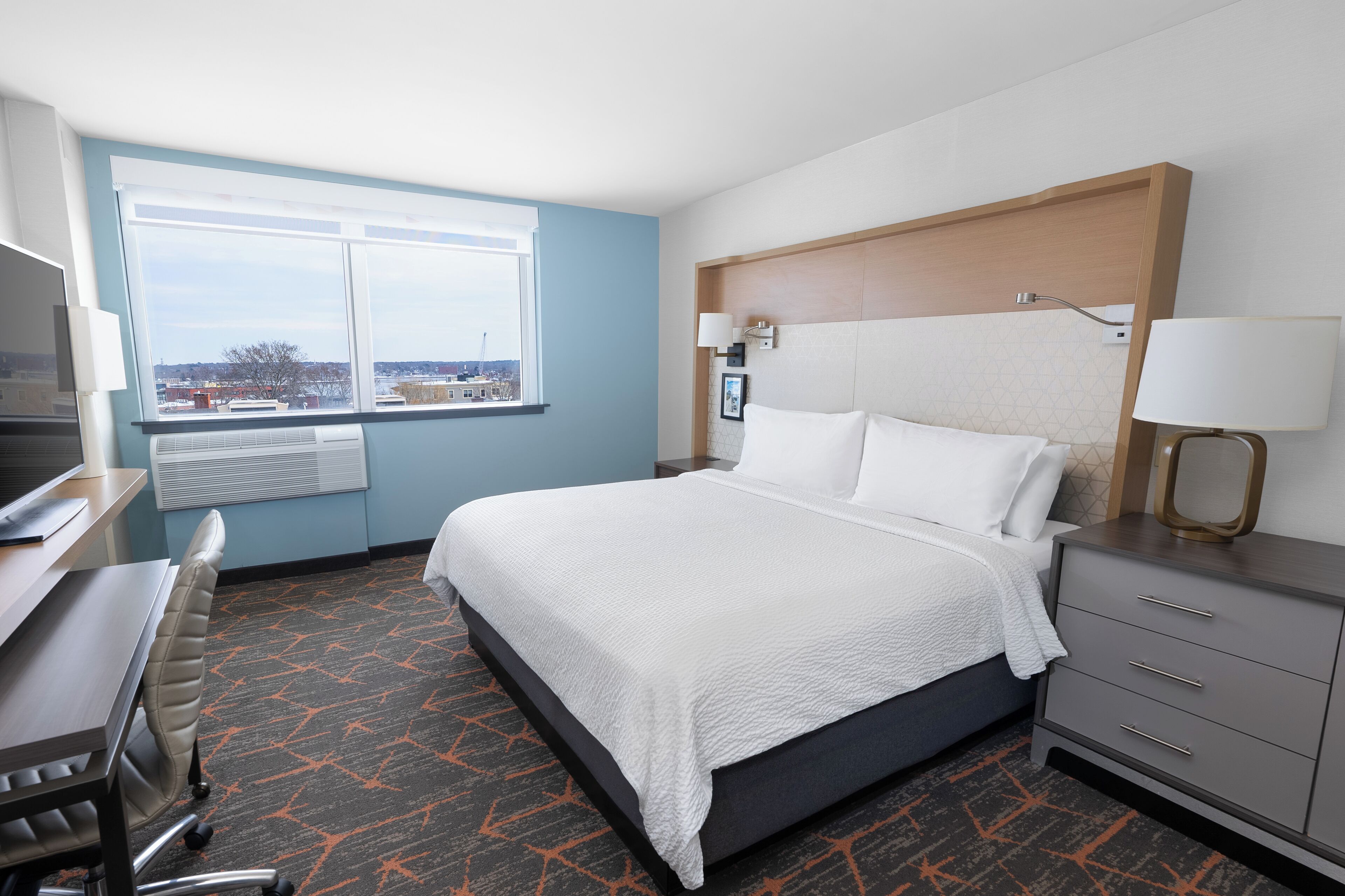 Quarto de hotel com cama, escrivaninha, cadeira, mesa de cabeceira, abajur e janela com vista para edifícios e árvores.
