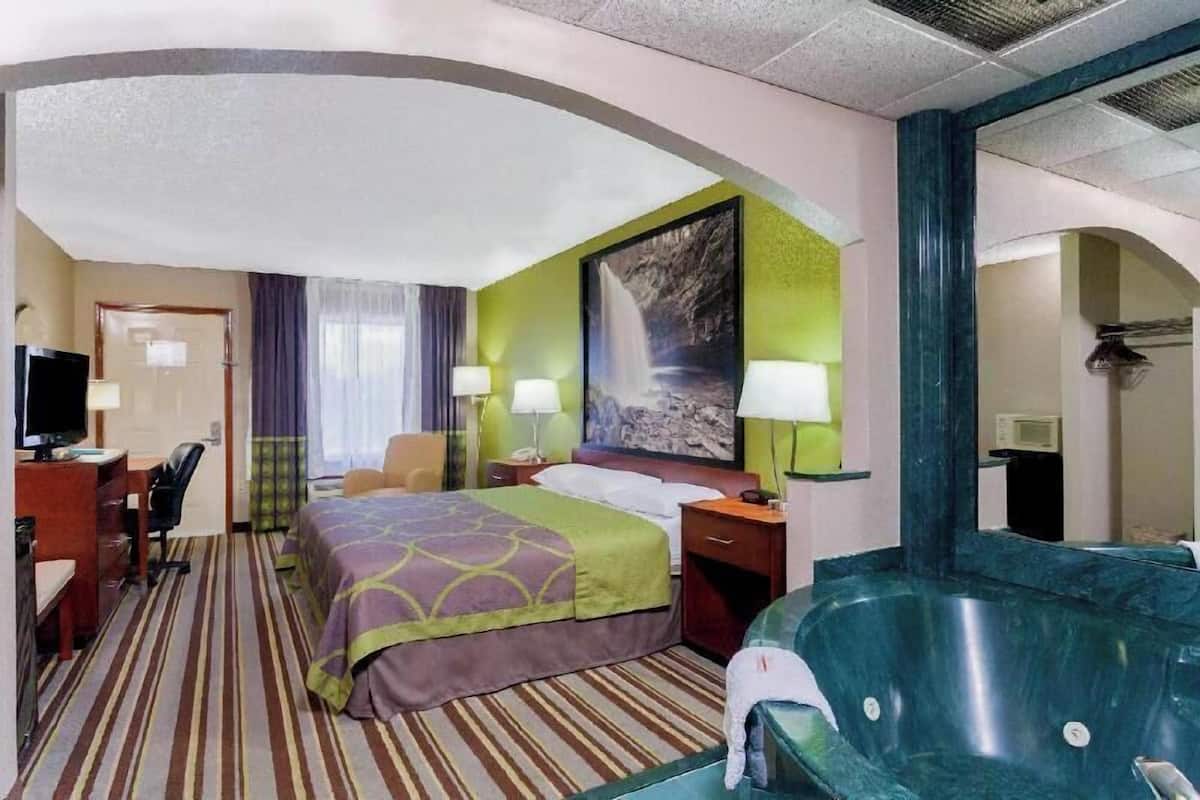 Quarto de hotel com cama, escrivaninha, cadeira, luminárias, um quadro grande, banheira e televisão.