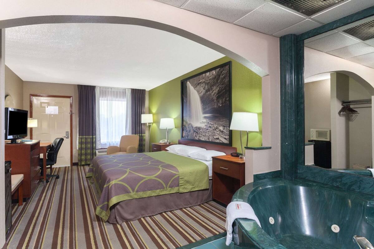 Quarto de hotel com cama, escrivaninha, cadeira, abajur, mesa de cabeceira, telefone, quadro, espelho grande e hidromassagem.
