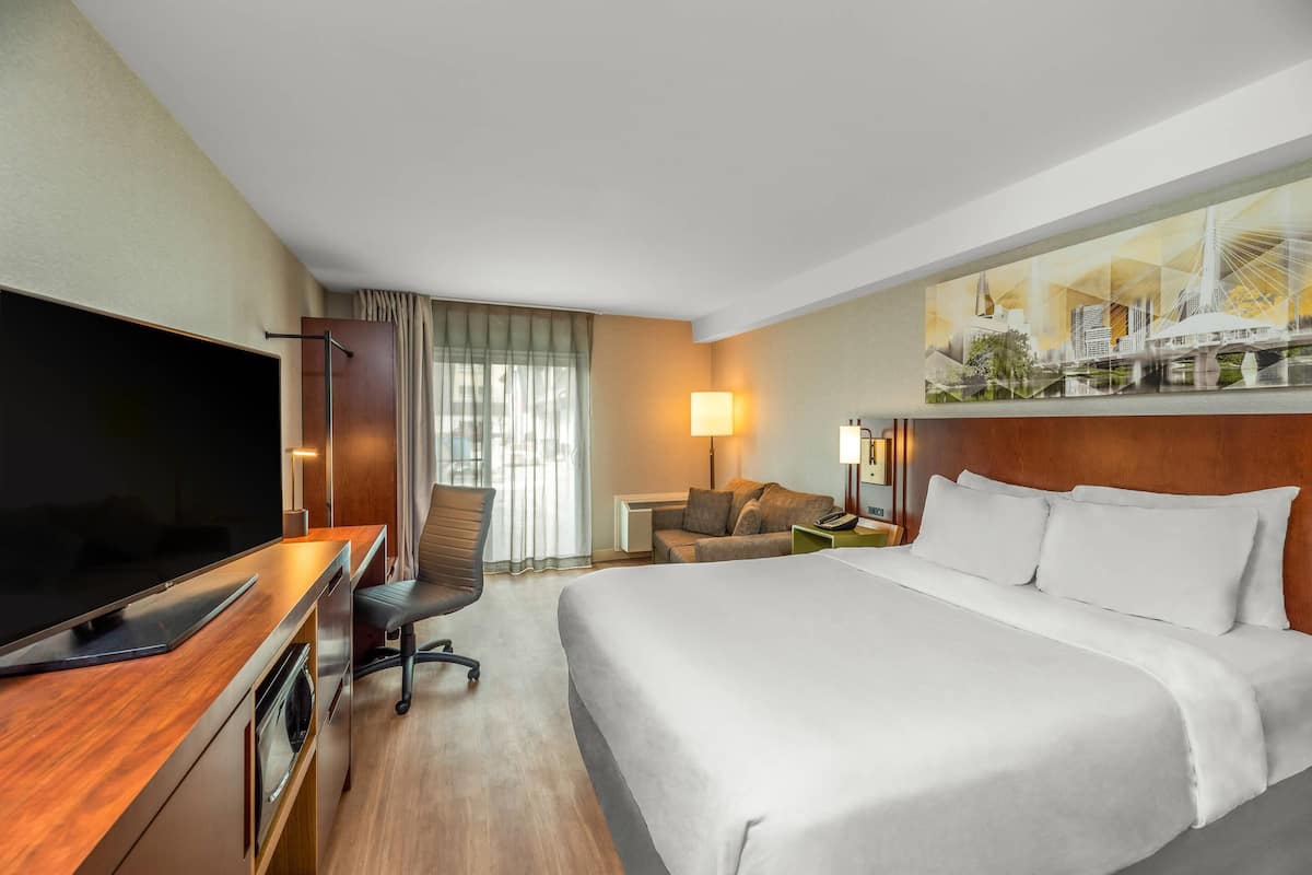 Quarto de hotel com uma cama grande, uma escrivaninha com televisão de tela plana, uma cadeira, um sofá e um quadro de uma ponte.