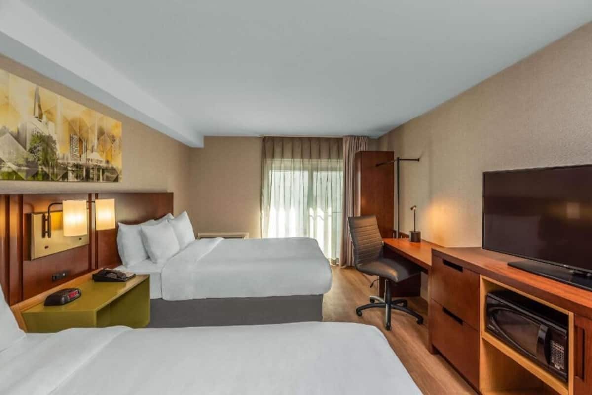 Quarto de hotel com duas camas, uma escrivaninha com televisão, uma cadeira, um abajur e um quadro na parede.