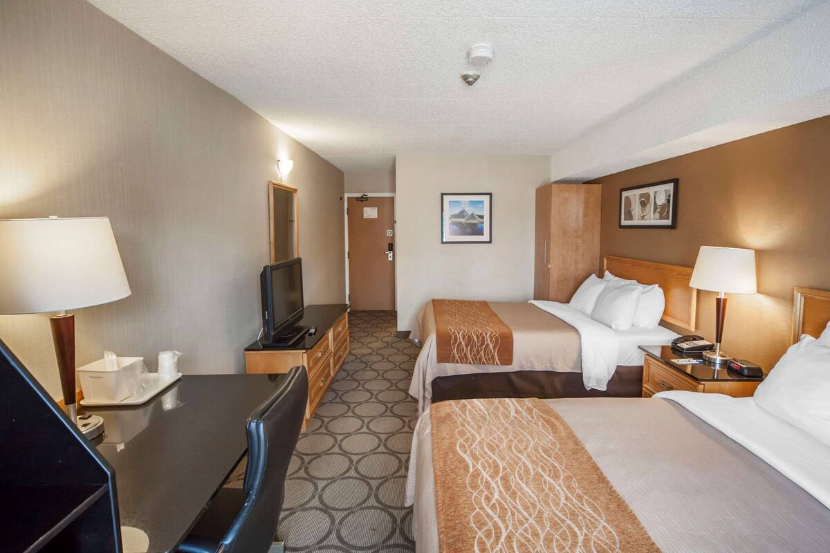 Quarto de hotel com duas camas, uma escrivaninha com televisão, uma cadeira e uma pequena mesa com abajur.