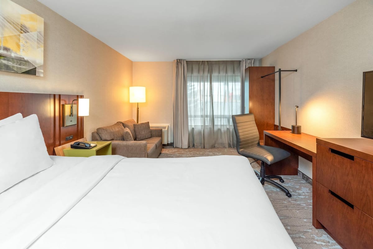 Quarto de hotel com uma cama grande, uma escrivaninha, uma cadeira, uma mesinha e uma luminária.