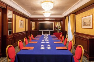Uma sala de conferências com uma mesa comprida, cadeiras vermelhas, um candelabro e quadros nas paredes.
