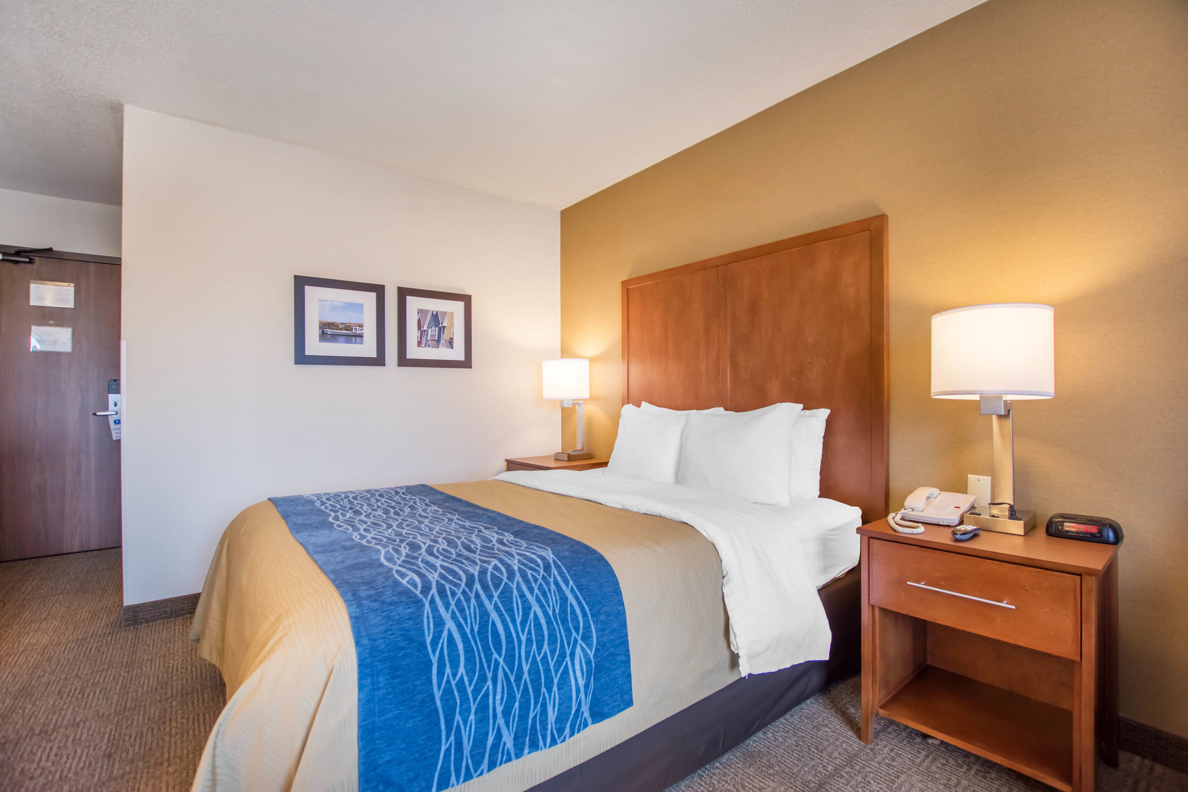 Quarto de hotel com uma cama, duas luminárias de cabeceira, um criado-mudo com telefone e controle remoto, e duas imagens emolduradas na parede.