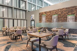 Espaço de refeições de hotel com mesas, cadeiras, parede de pedra e uma grande janela com cortinas.