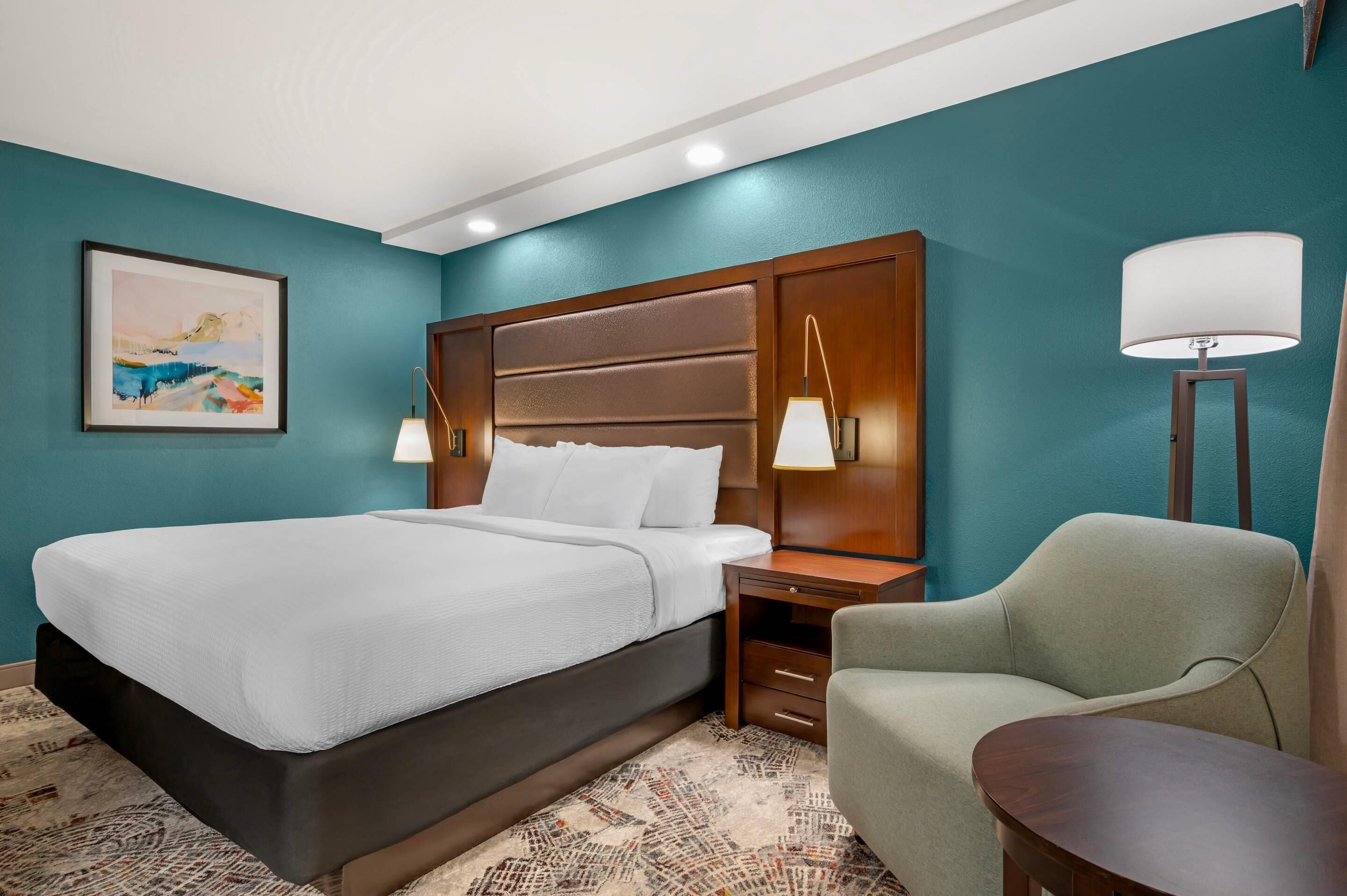 Quarto de hotel com uma cama grande, uma mesa de cabeceira, uma cadeira e uma luminária.