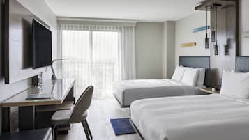 Quarto de hotel com duas camas, uma escrivaninha, uma cadeira, uma televisão e uma janela com cortinas leves.